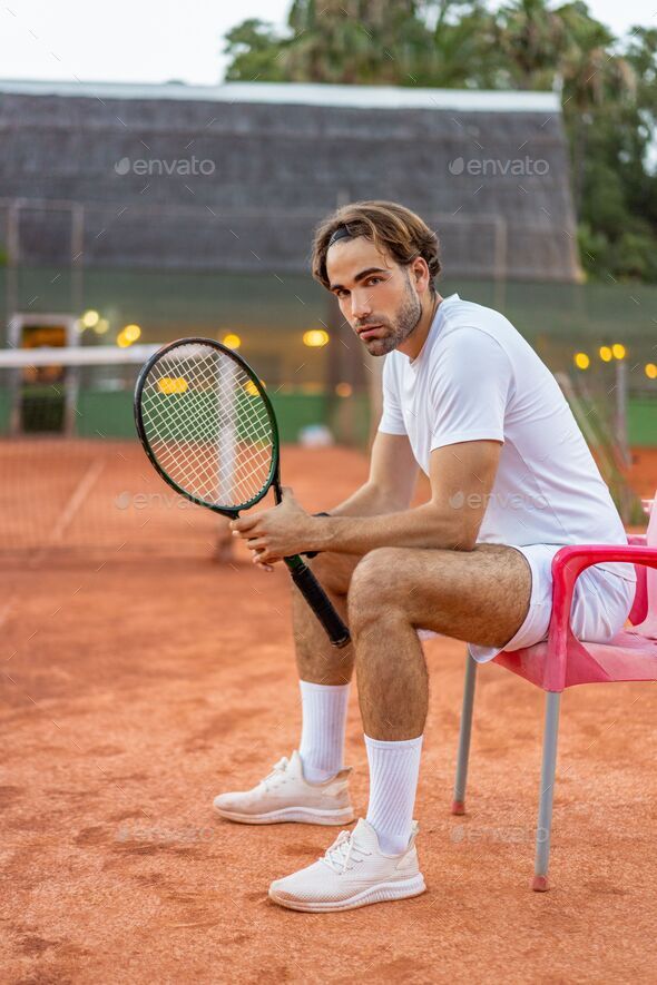 Caucasian young man sitting on a chair holding tennis racket on a dirt ...