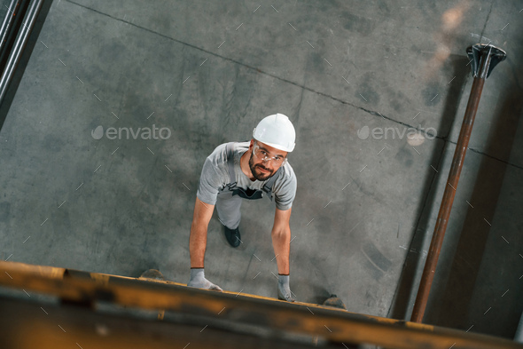 Going up on the ladder, top view. Young factory worker in grey uniform ...