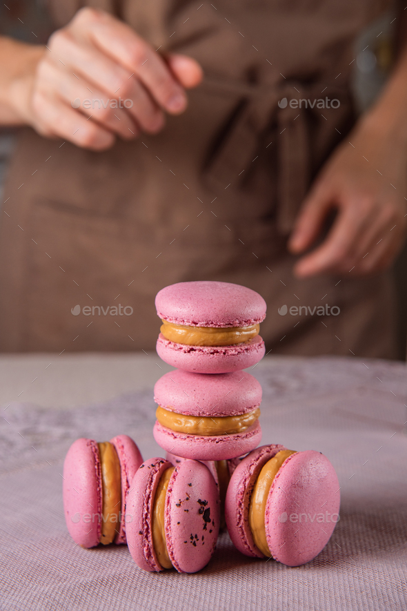 pink macaroon desert stacked in column on table on woman in apron background. cook sweets at ...