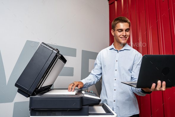 Positive young man using printer in the modern office Stock Photo by ...