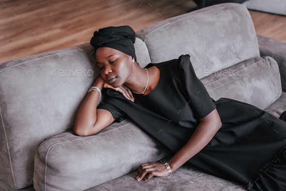 Exhausted African girl in black turban and black dress laying on couch ...