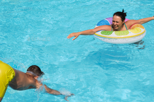 Boy swims toward a colorful float, mother reaches out; a lively pool ...
