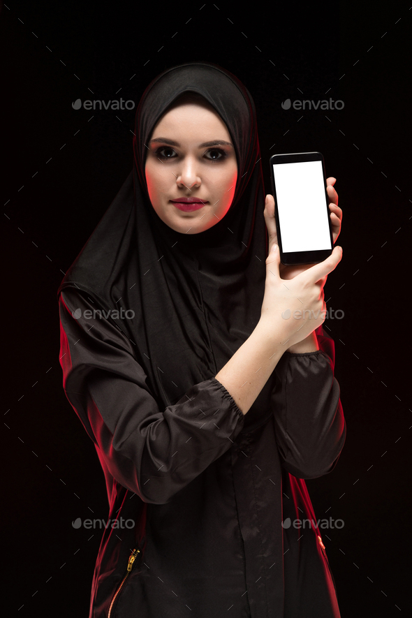 Portrait of beautiful smart young muslim woman wearing black hijab advertising mobile phone in ...