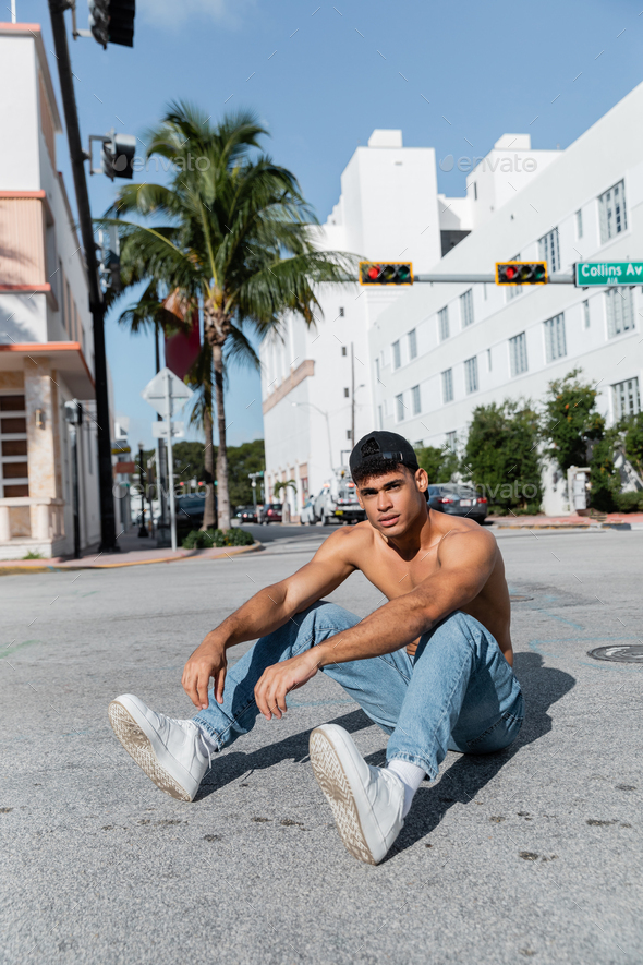 muscular cuban man in baseball cap and blue jeans sitting on road in ...