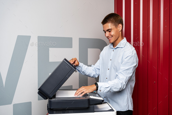 Positive young man using printer in the modern office Stock Photo by ...