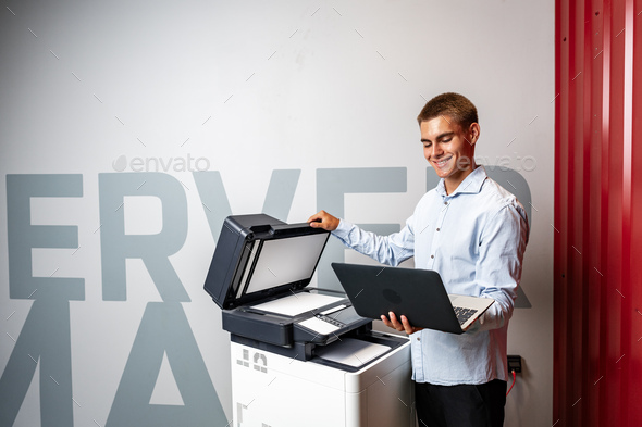 Positive young man using printer in the modern office Stock Photo by ...