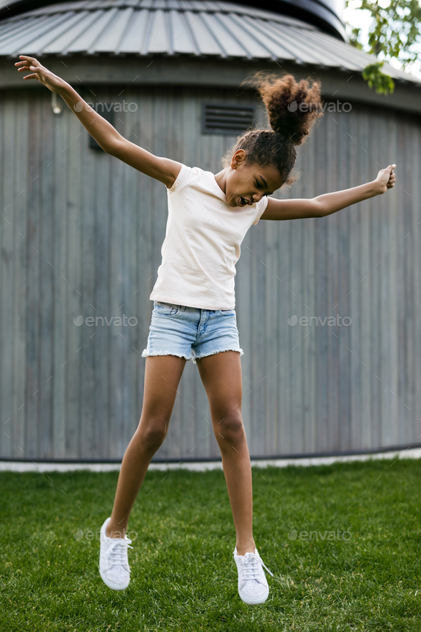 Cheerful girl jumping on grass at backyard Stock Photo by ArtemVarnitsin