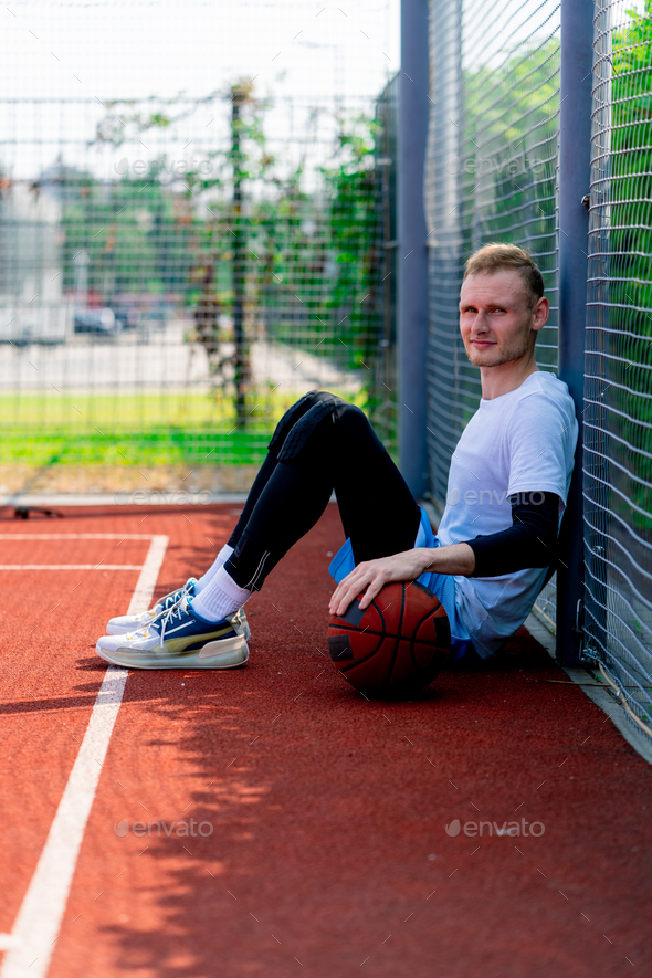 A tall guy basketball player with a ball in his hand sits leaning on ...