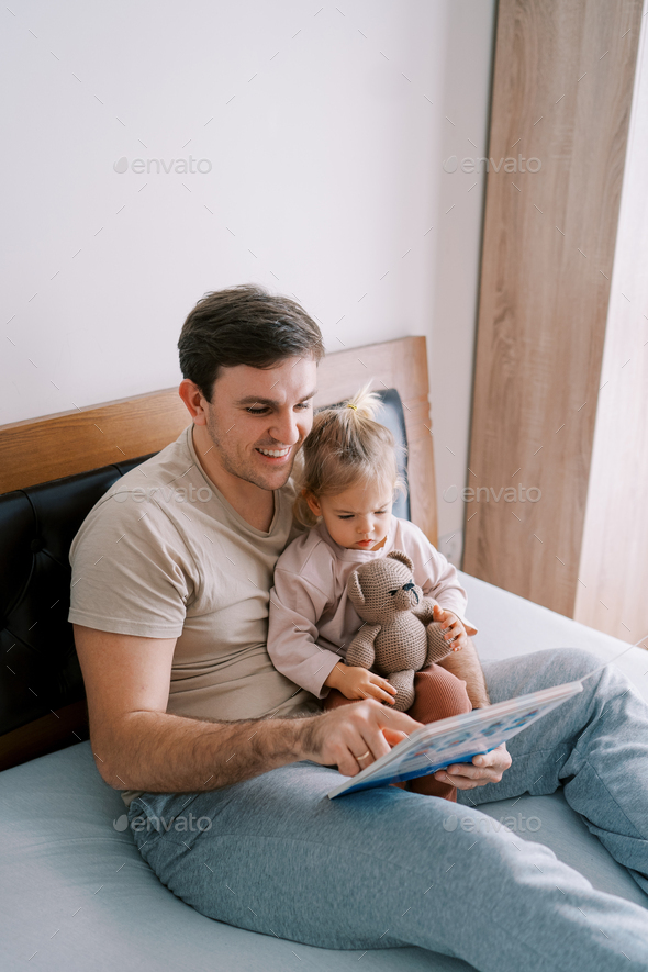Smiling dad reading a book to a little girl sitting with a teddy bear on his lap on the bed ...