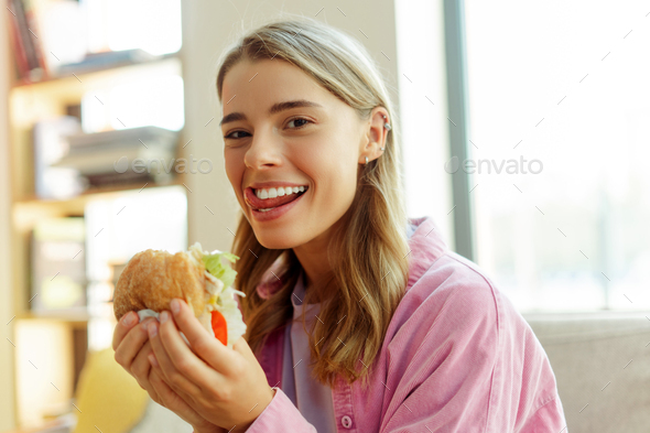 Hungry smiling woman lick lips eating burger looking at camera sitting ...