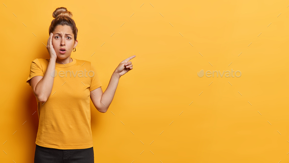 Photo of embarrassed shocked young woman keeps hand on cheek points ...