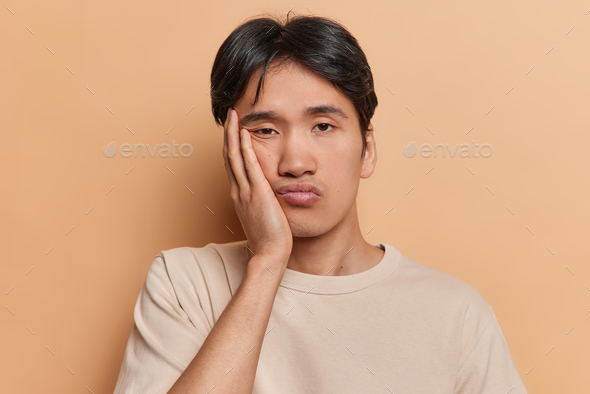 Portrait of displeased Japanese man has dull day keeps hand on cheek ...