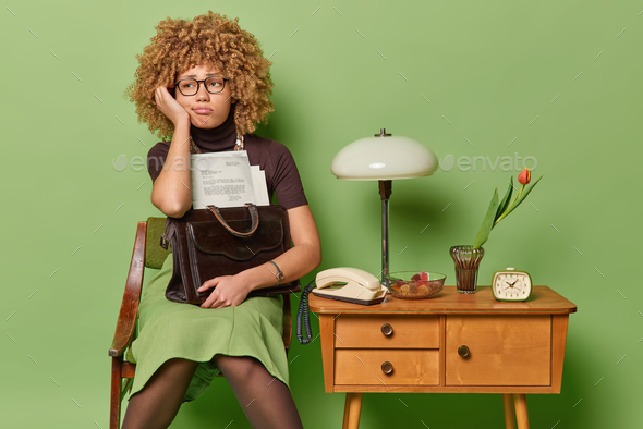 Indoor shot of thoughtful curly haired lady poses with briefcase full ...