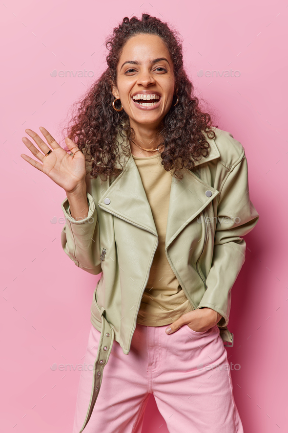 Attractive joyful Brazilian woman keeps hand in pocket waves hello with ...