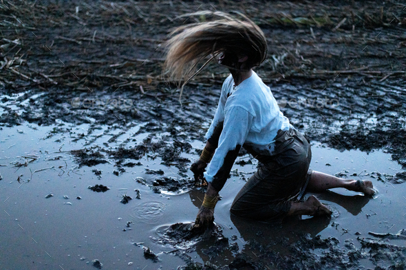 Dramatic portrait of a young woman kneeling in the black mud on field ...