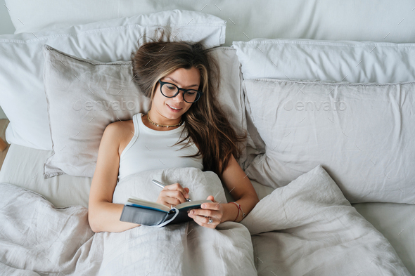 Relaxed young woman writing in a journal while lying in bed, enjoying ...