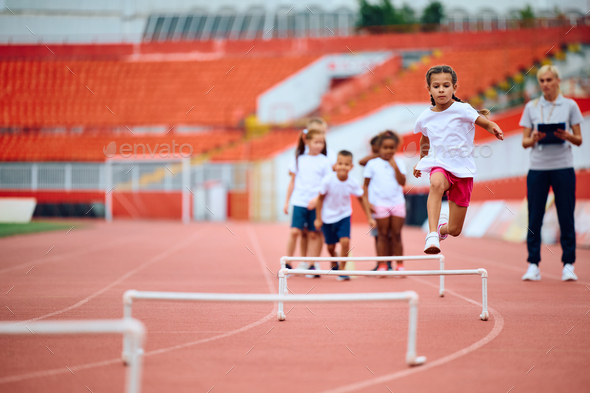 Little girl jumping over hurdle during PE class at the stadium. Stock ...