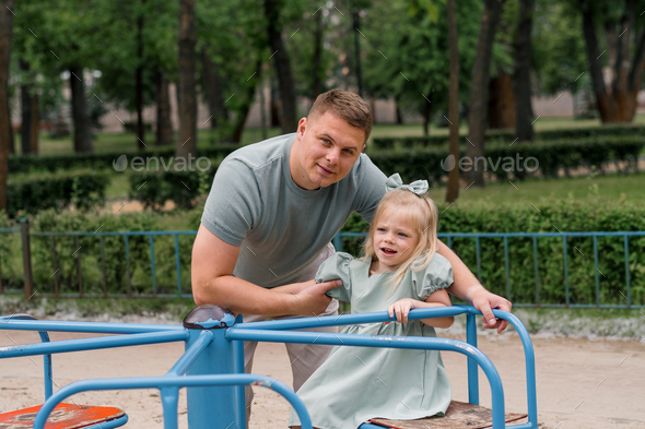 Little cute girl in green summer dress with bow on head on playground dad rides daughter on ...