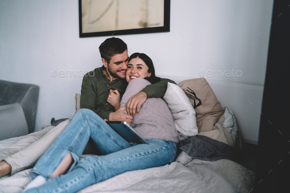 Happy couple sitting on bed together and cuddling while having laptops ...