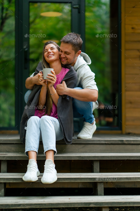 A thoughtful woman sits on the doorstep of a wooden house with a cup of ...