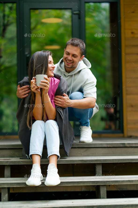 A thoughtful woman sits on the doorstep of a wooden house with a cup of ...