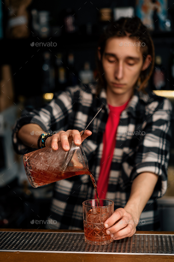 bartender pouring negroni alcoholic cocktail with crystal ice cubes from measuring glass into ...