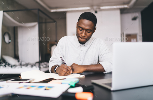 Focused young African American man writing in notebook while working on ...
