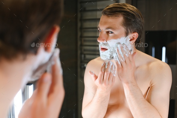 Young smiling Caucasian shirtless man applying shaving foam on face in front of mirror Stock ...