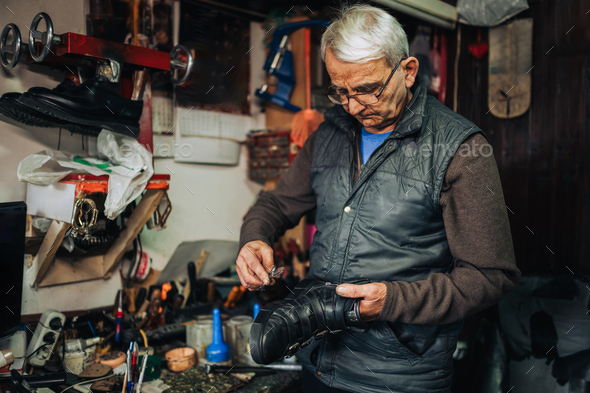 An old shoemaker is standing with boots in his hands and repairing them ...