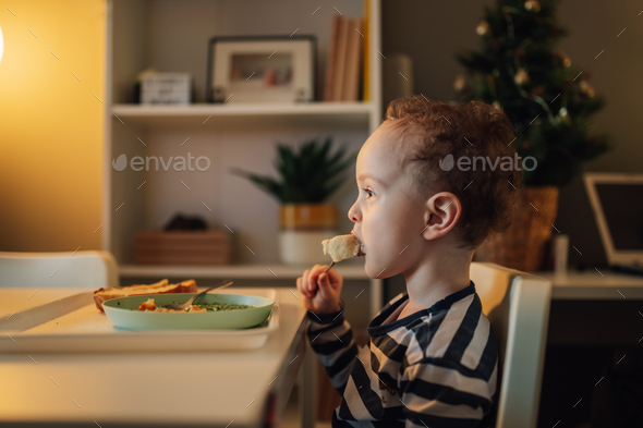 Side view of a hungry toddler sitting at dining table at home and ...
