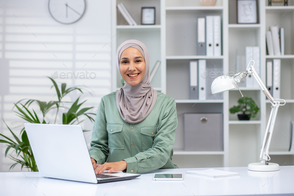 Professional muslim woman with hijab smiling while working on laptop in ...