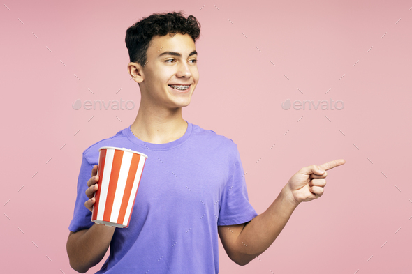 Handsome, smiling boy with braces holding bucket of popcorn pointing ...