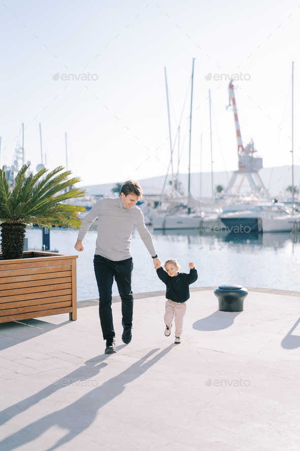 Smiling dad and little girl walk along the pier holding hands and jumping Stock Photo by Nadtochii