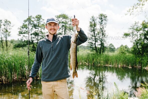 Happy cheerful young fisherman hold a big fish pike on a background of ...