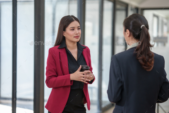 Two women in business attire are talking in a room with glass walls ...