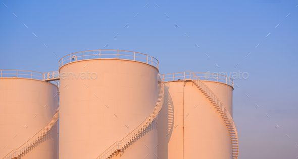 Orange sunlight on surface of 3 storage fuel tanks against blue evening ...