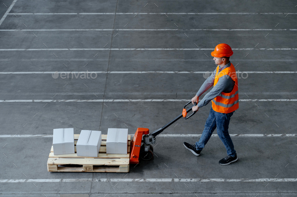 Worker wearing protective helmet, work wear, vest, carrying pallet ...