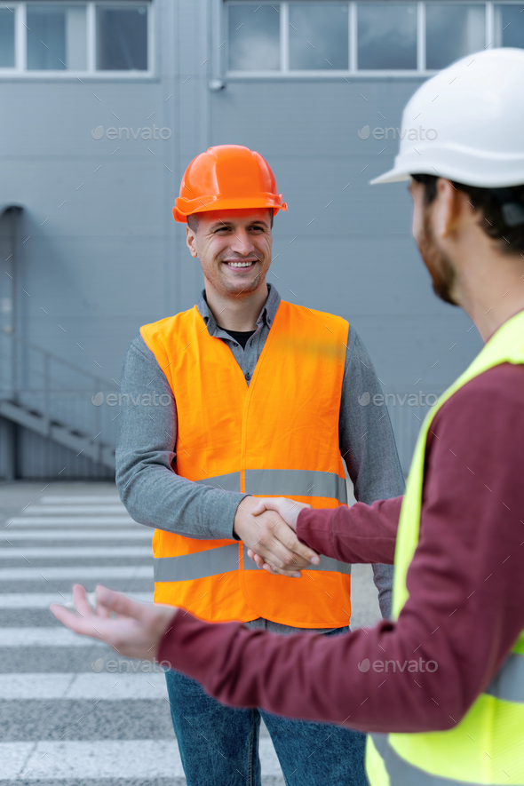Handsome, smiling men, workers wearing hard hats, vests shaking hands ...