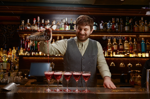 Smiling bartender pouring freshly prepared cocktail into several ...