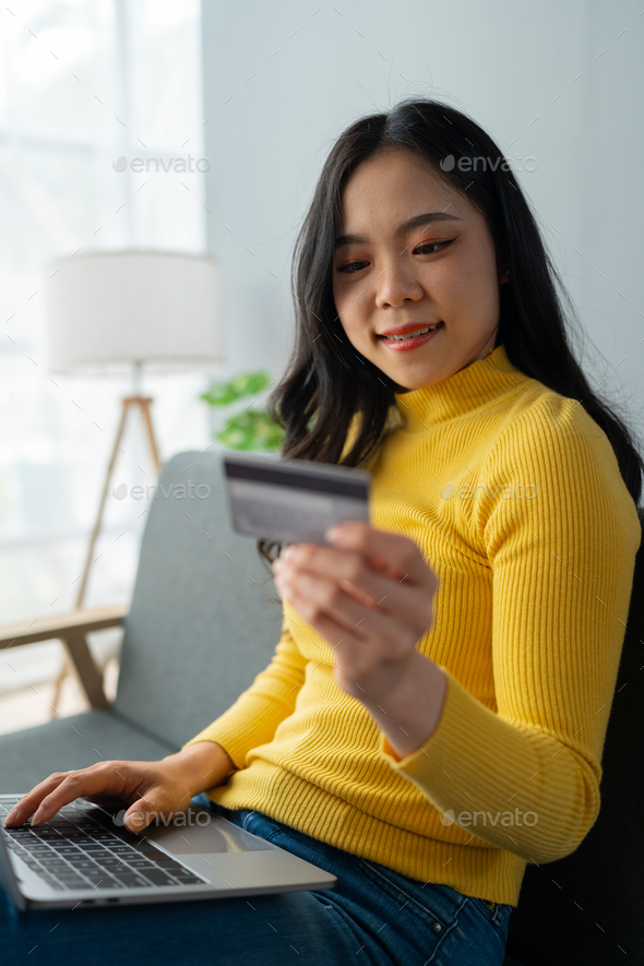 Young Asian woman holding credit card and smartphone sitting on sofa at ...