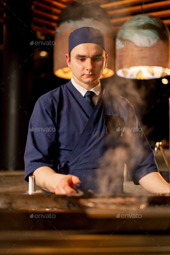 in a Japanese restaurant chef in a blue uniform fries fish on the grill ...