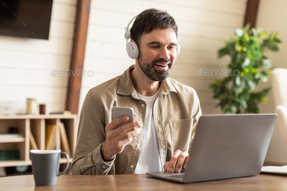 Smiling Man With Headphones Using Smartphone While Working on Laptop ...
