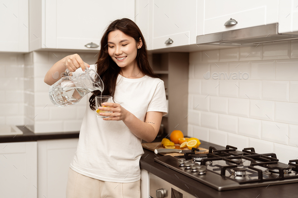 Happy Young Woman Pouring Water Into Glass In Modern Kitchen Stock Photo by puhhha