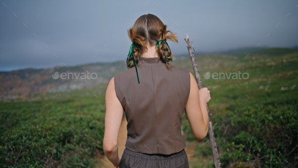 Woman journeying green hill with stick alone. Rear view serene girl ...