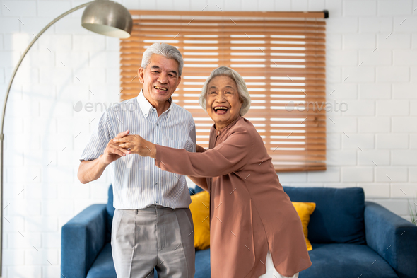 Portrait of Asian senior couple dance together in living room at home. Stock Photo by s_kawee