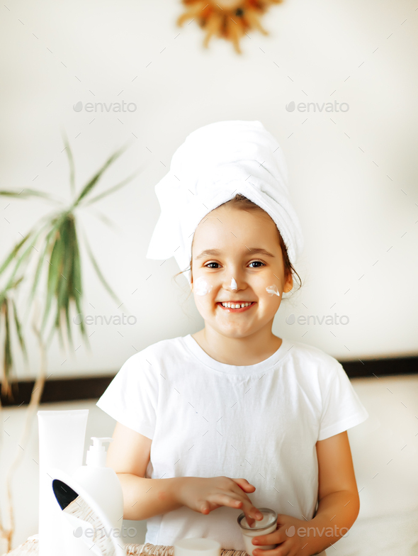 Funny little girl with towel on her head uses facial cream. Spa ...