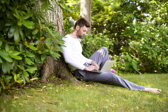 Young man sitting against tree, using laptop Stock Photo by Image-Source