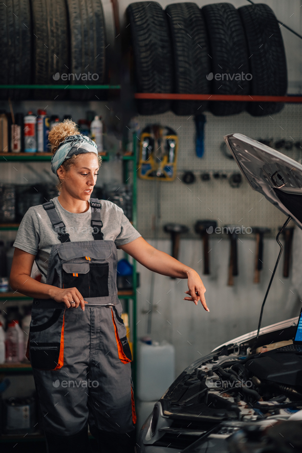 Female mechanic pointing under broken car hood at mechanic workshop ...