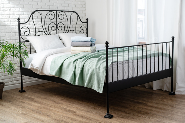 Black Metal Bed Frame With White Brick Wall And Green Blanket Stock Photo by FabrikaPhoto
