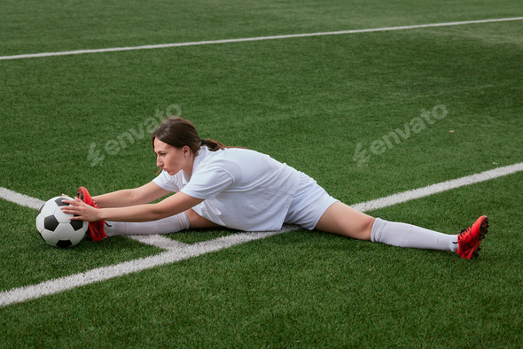 A young female soccer player stretches on a soccer field and reaches ...